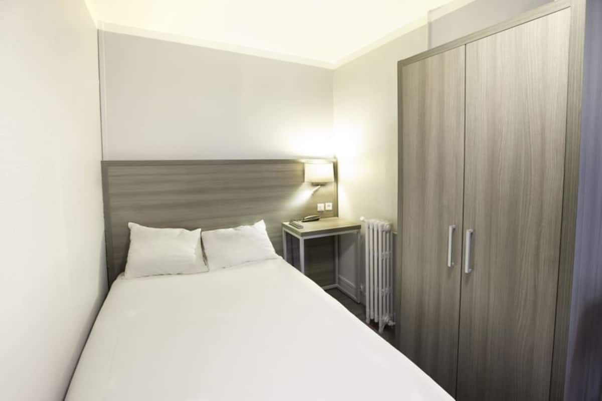 Quarto de hotel com cama, mesa de cabeceira, abajur e guarda-roupa.