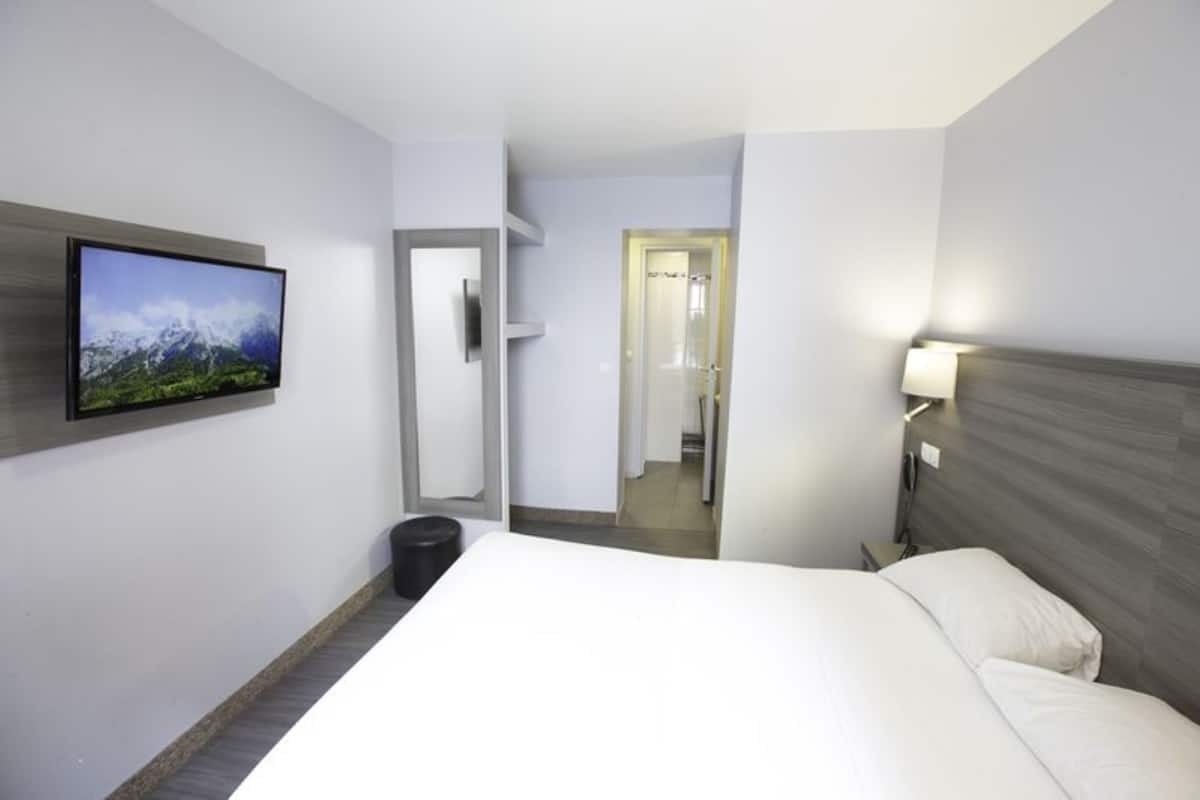 Quarto de hotel com uma cama, televisão presa na parede, mesa de cabeceira e porta para uma casa de banho.
