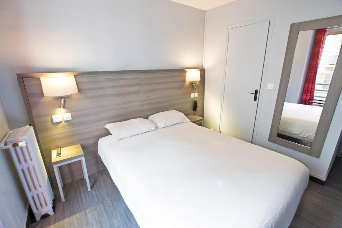 Quarto de hotel com uma cama grande, luminárias de cabeceira, mesa de cabeceira, radiador e espelho.