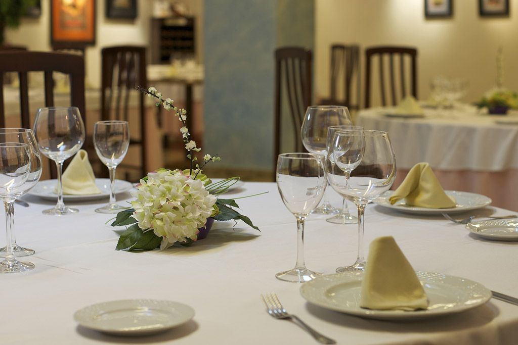 Mesa de jantar posta com pratos brancos, copos de vinho e um arranjo central de flores brancas.