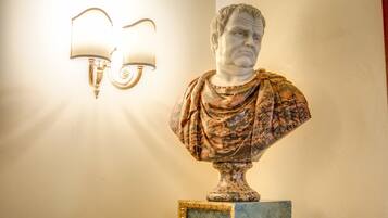 Escultura em busto sobre pedestal com base de mármore, posicionada em frente a uma parede com duas luminárias de parede.