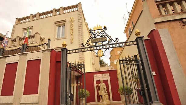 Entrada de hotel com uma estátua, uma placa com o texto 'Aurelius Hotel' e um portão decorativo.