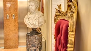 Busto de mármore sobre pedestal em corredor com paredes de mármore e espelho dourado ornamentado.