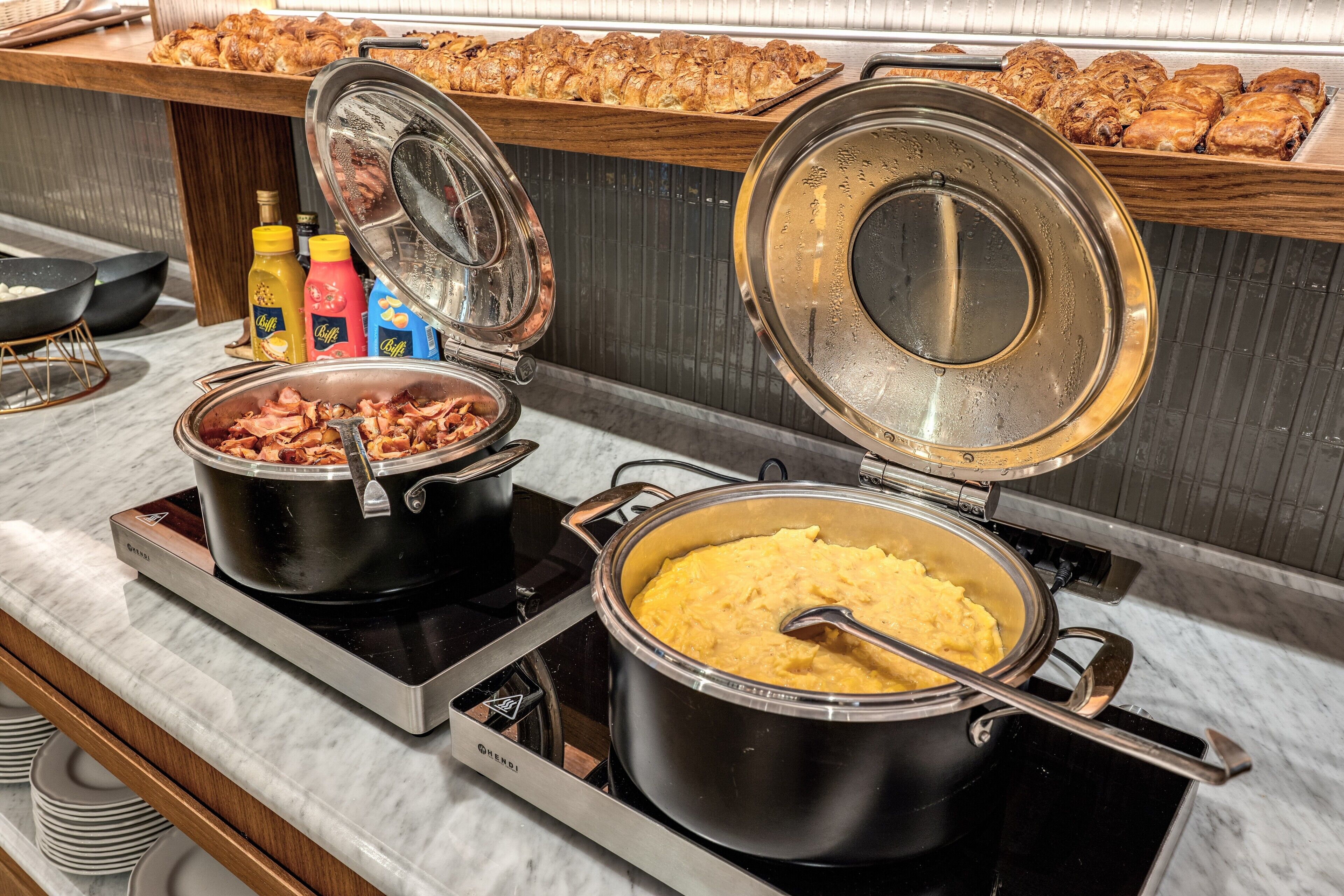 Buffet com diversos alimentos, incluindo um recipiente de purê de batata e uma embalagem de bacon.