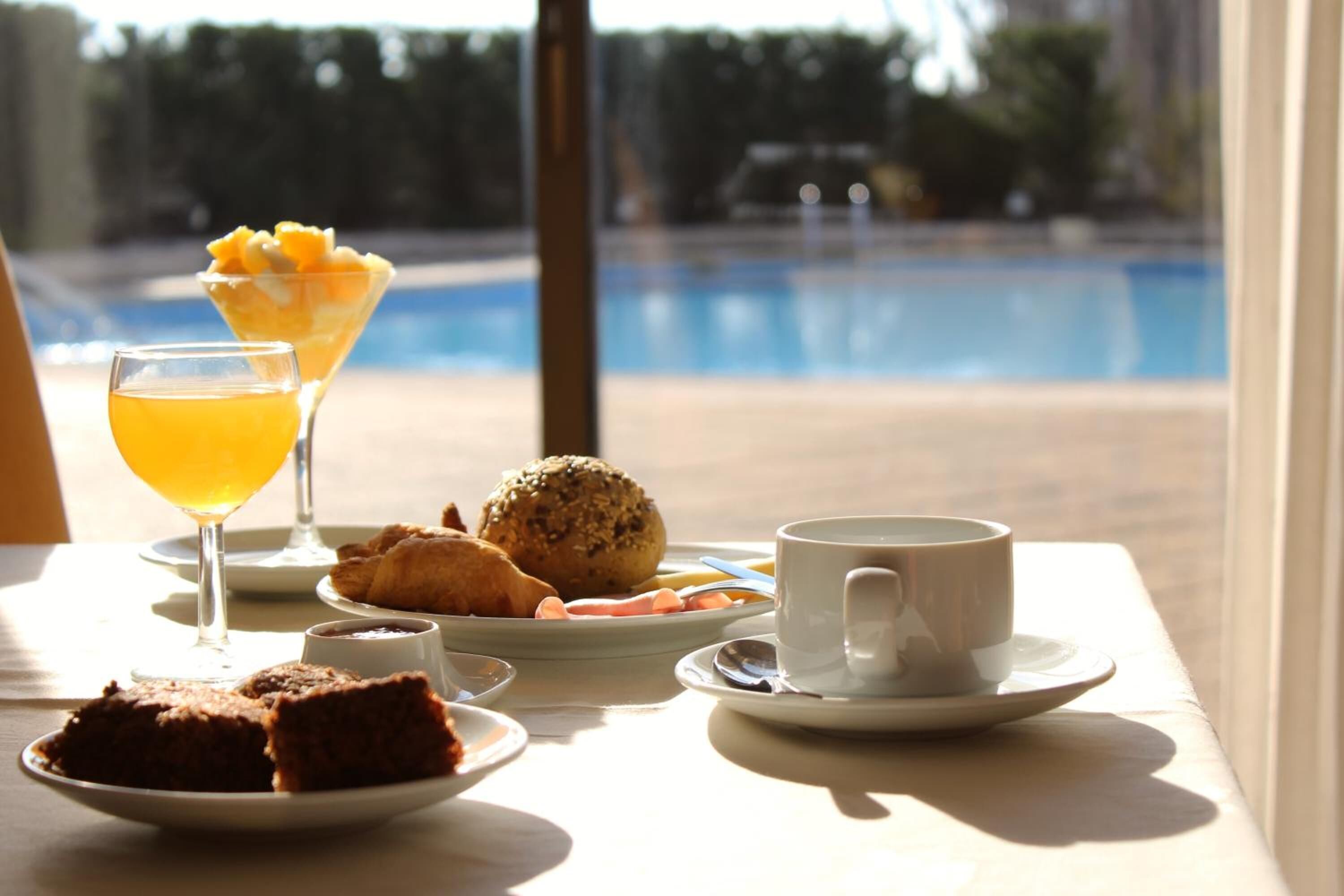 Mesa de café da manhã com copo de suco de laranja, xícara de café, pães e uma tigela de frutas à beira da piscina.