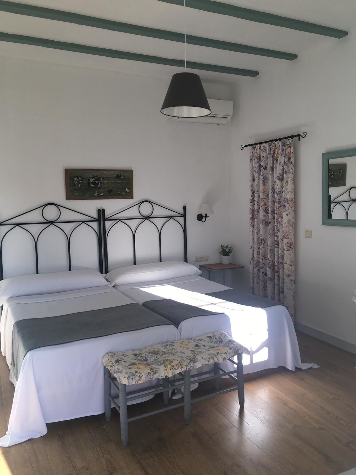 Quarto com duas camas, uma luminária pendente, cortina florida, uma mesinha e um espelho fixado na parede.