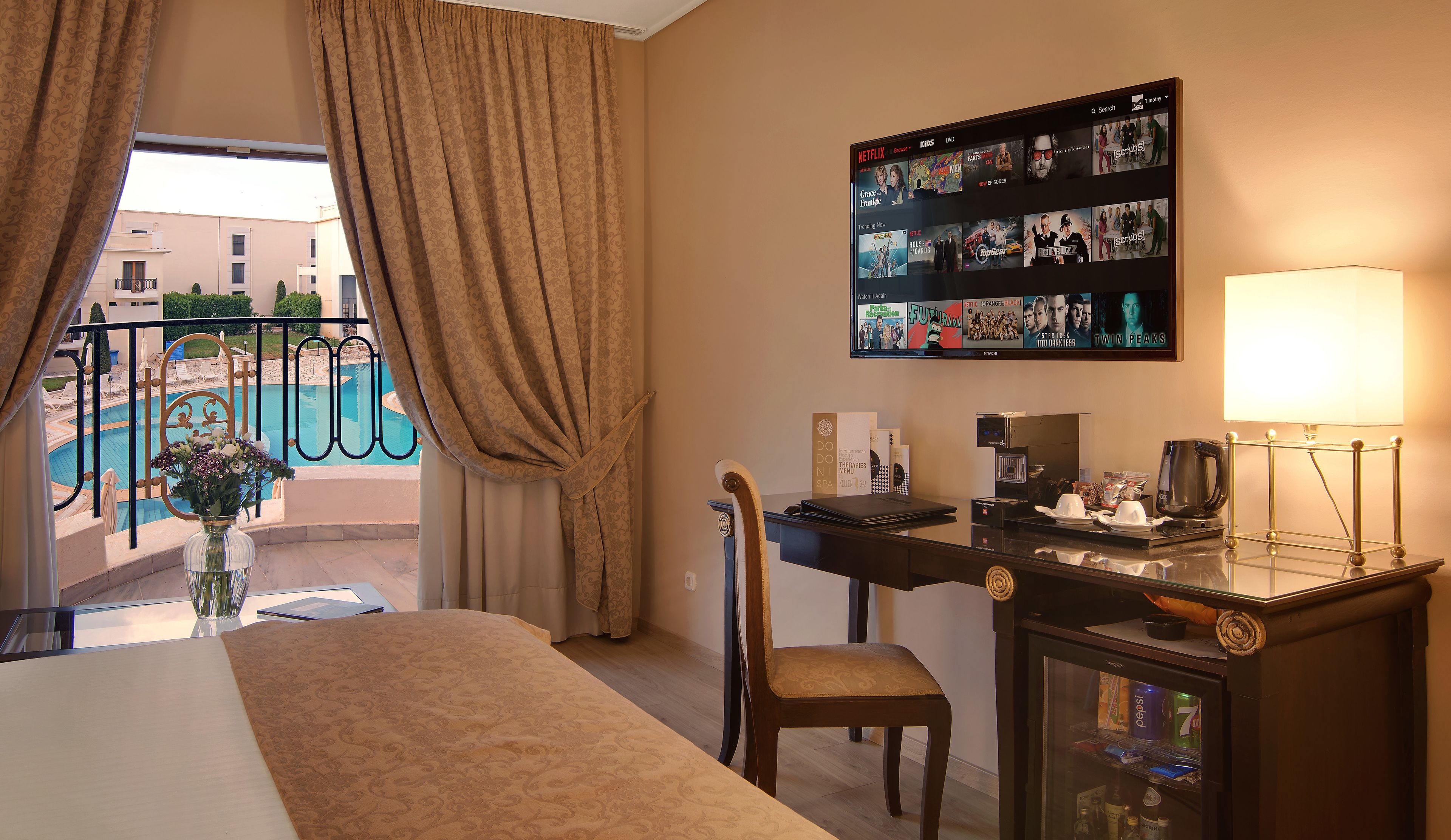Quarto de hotel com cama, escrivaninha com luminária, cadeira, TV exibindo Netflix e vista para uma piscina.