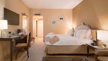 Quarto de hotel com cama, mesa de cabeceira, escrivaninha, cadeira e televisão.