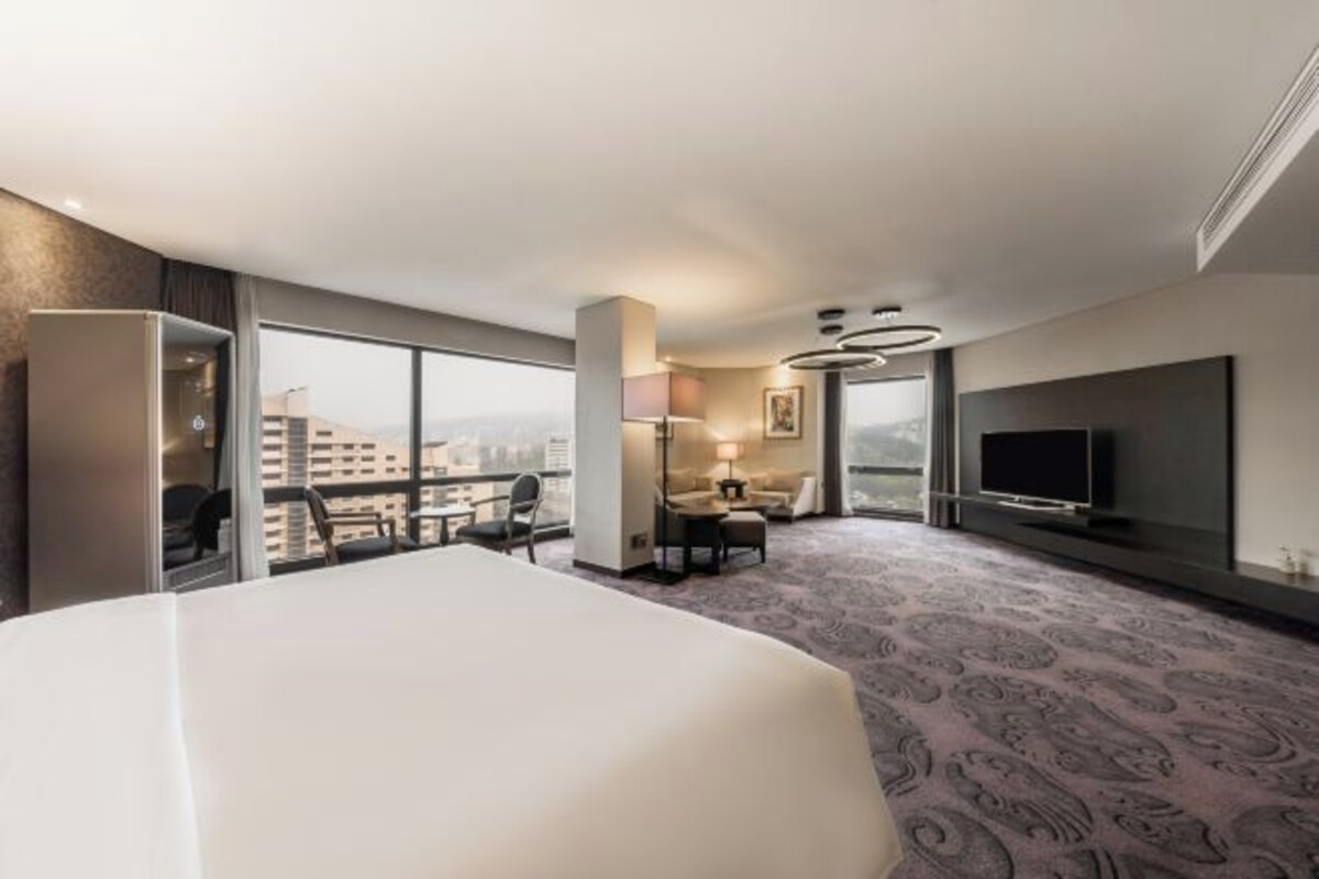 Quarto de hotel com uma cama grande, uma escrivaninha, uma cadeira, uma televisão e vista para a cidade.