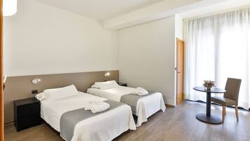 Quarto de hotel com duas camas, uma mesa pequena com uma cadeira e uma janela com cortinas.