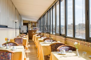 Um restaurante com mesas e cadeiras dispostas em fileiras, janelas amplas e vista para edifícios do lado de fora.