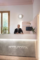 Balcão de recepção de hotel com um homem sentado atrás, uma televisão na parede e plantas em vasos.