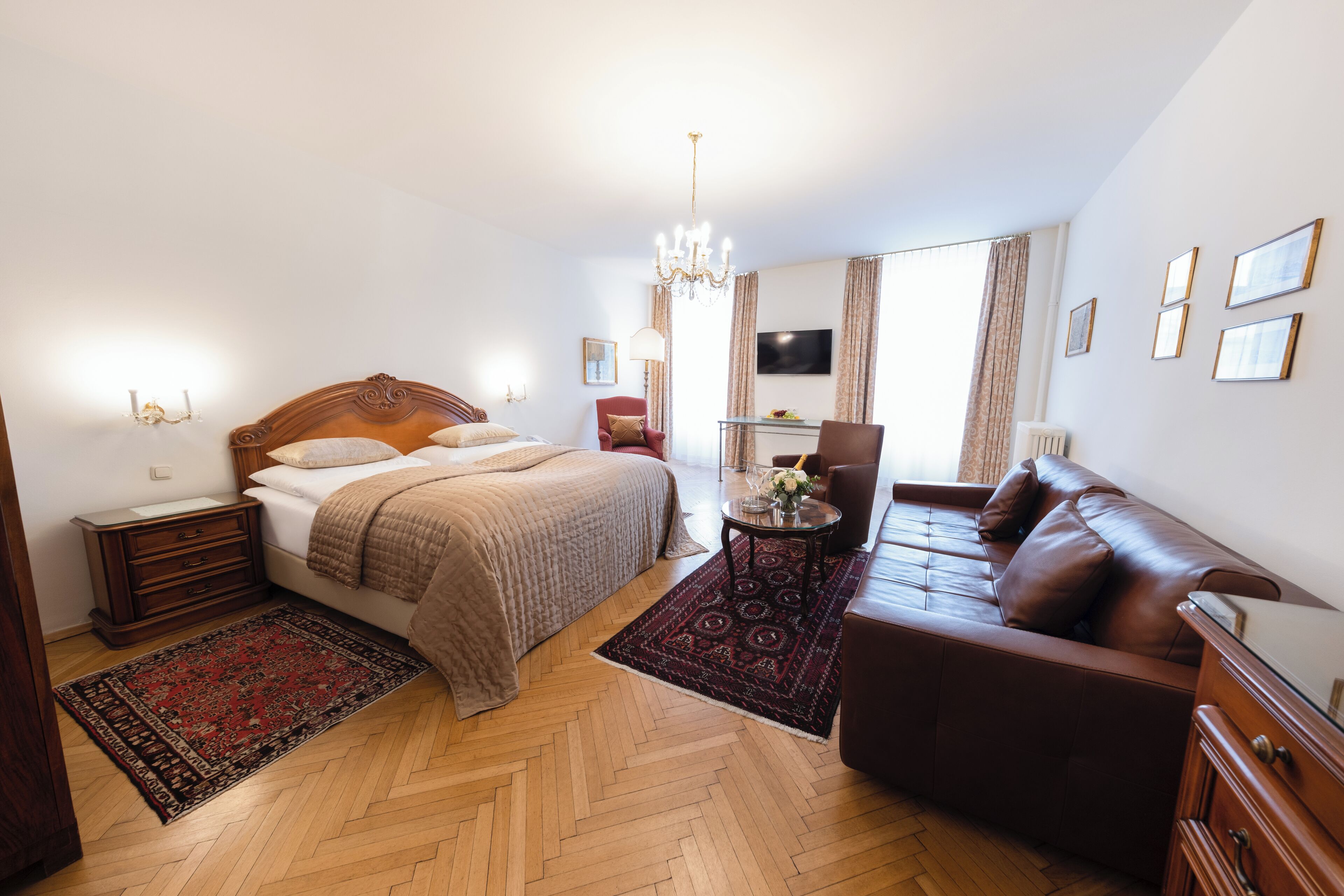 Um quarto com cama, mesa de cabeceira, cômoda, sofá, mesa com um vaso e televisão na parede.