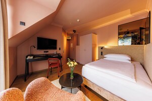 Quarto de hotel com cama, escrivaninha com cadeira, mesa pequena com um vaso de flores e televisão na parede.