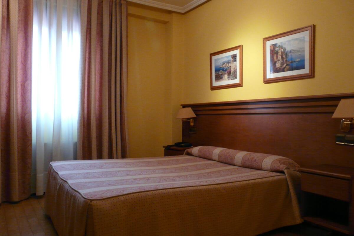 Quarto de hotel com cama, cortinas, mesa de cabeceira, abajur e duas fotos emolduradas na parede.