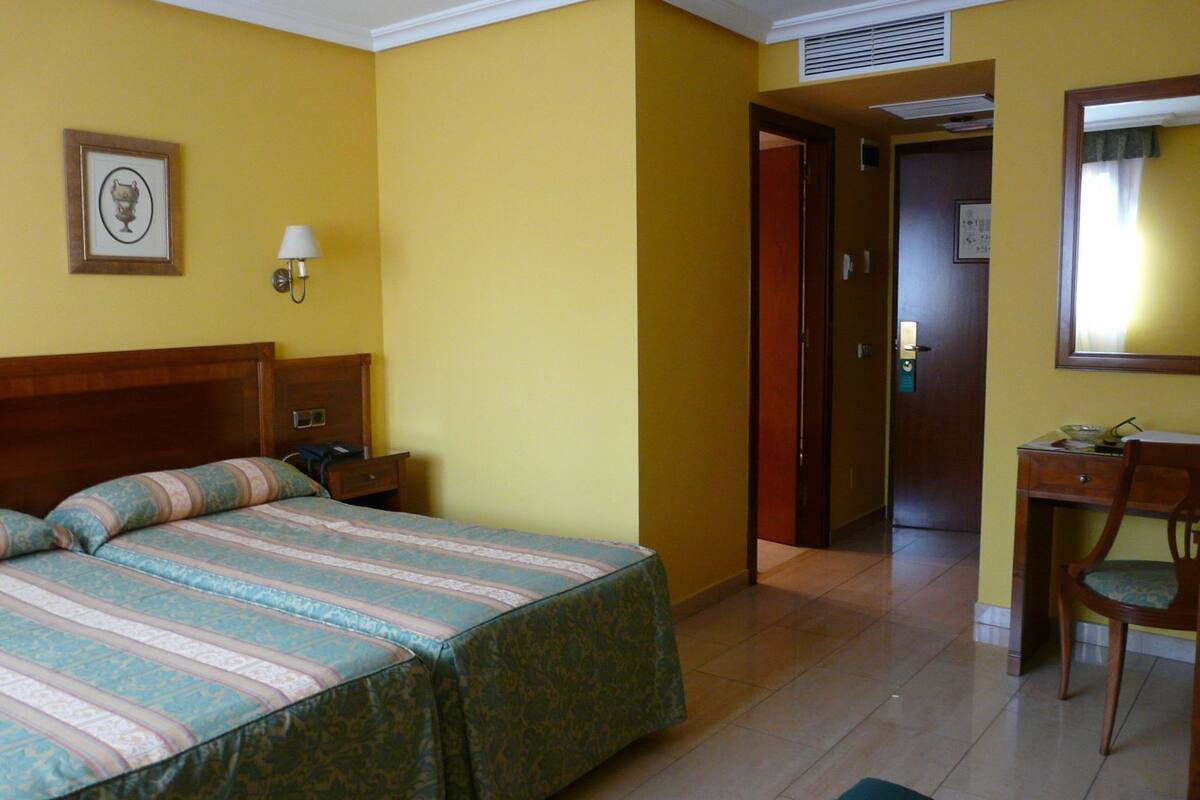 Quarto de hotel com uma cama, uma escrivaninha, uma cadeira, um criado-mudo, um abajur e uma porta que dá para outro quarto.