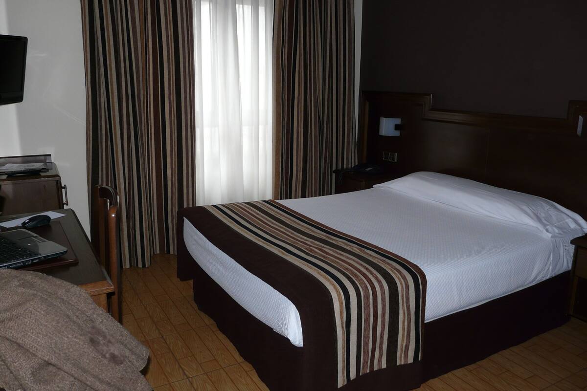 Quarto de hotel com cama, escrivaninha com laptop, cadeira, televisão e cortinas.