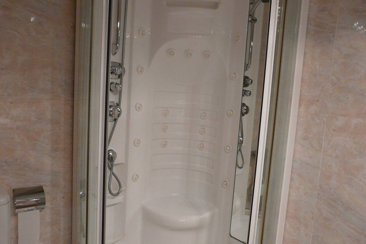 Box de chuveiro envidraçado com painel de controle na parede, vaso sanitário e dispensador de toalhas.