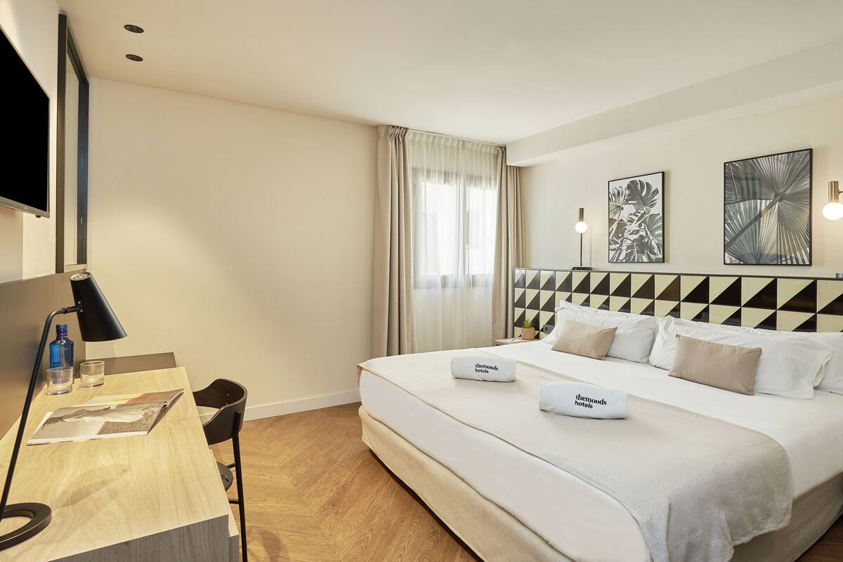 Quarto de hotel com uma cama, escrivaninha, cadeira, abajur e duas obras de arte na parede.