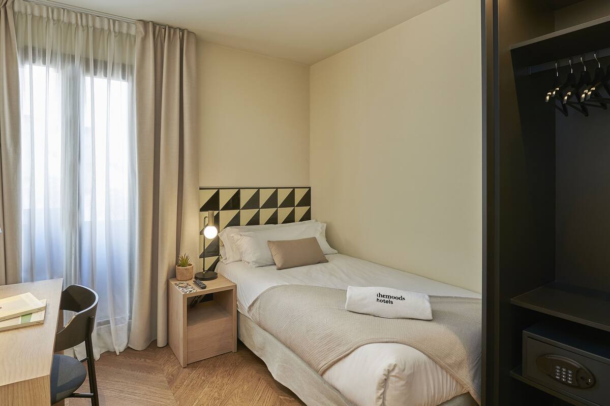 Quarto de hotel com uma cama, uma escrivaninha com abajur, uma cadeira, uma mesa de cabeceira com abajur e uma televisão.