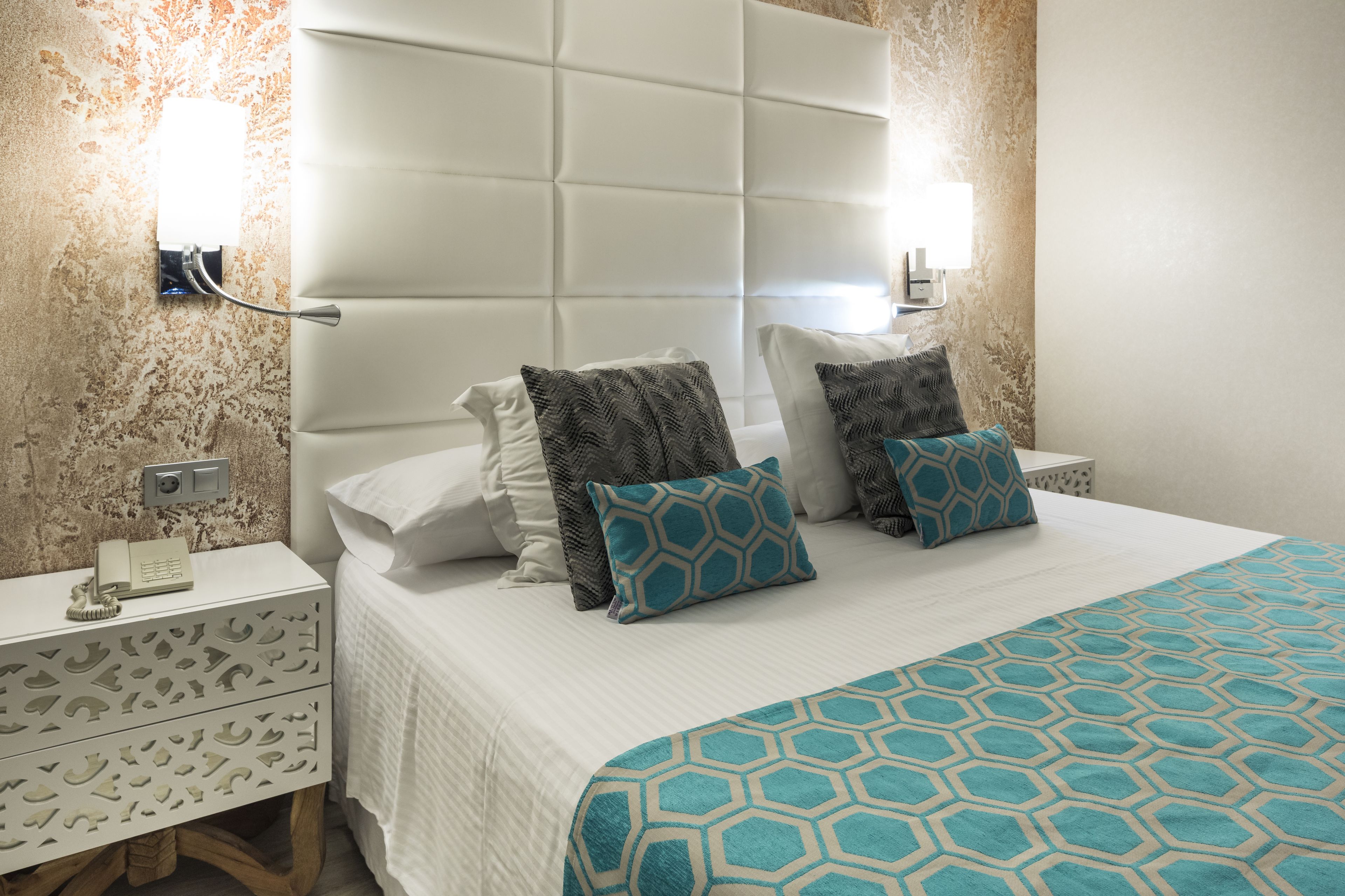 Uma cama bem arrumada com colcha estampada, duas almofadas decorativas, uma mesa de cabeceira com telefone e luminárias fixadas na parede.