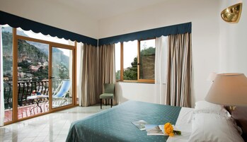 Quarto de hotel com cama, cadeira, varanda com mesa e cadeiras, e vista para uma paisagem montanhosa.