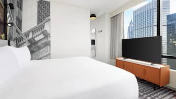 Quarto de hotel com uma cama grande, uma TV sobre um móvel de madeira, vista para a cidade e um painel com o letreiro 'Black Exchange'.