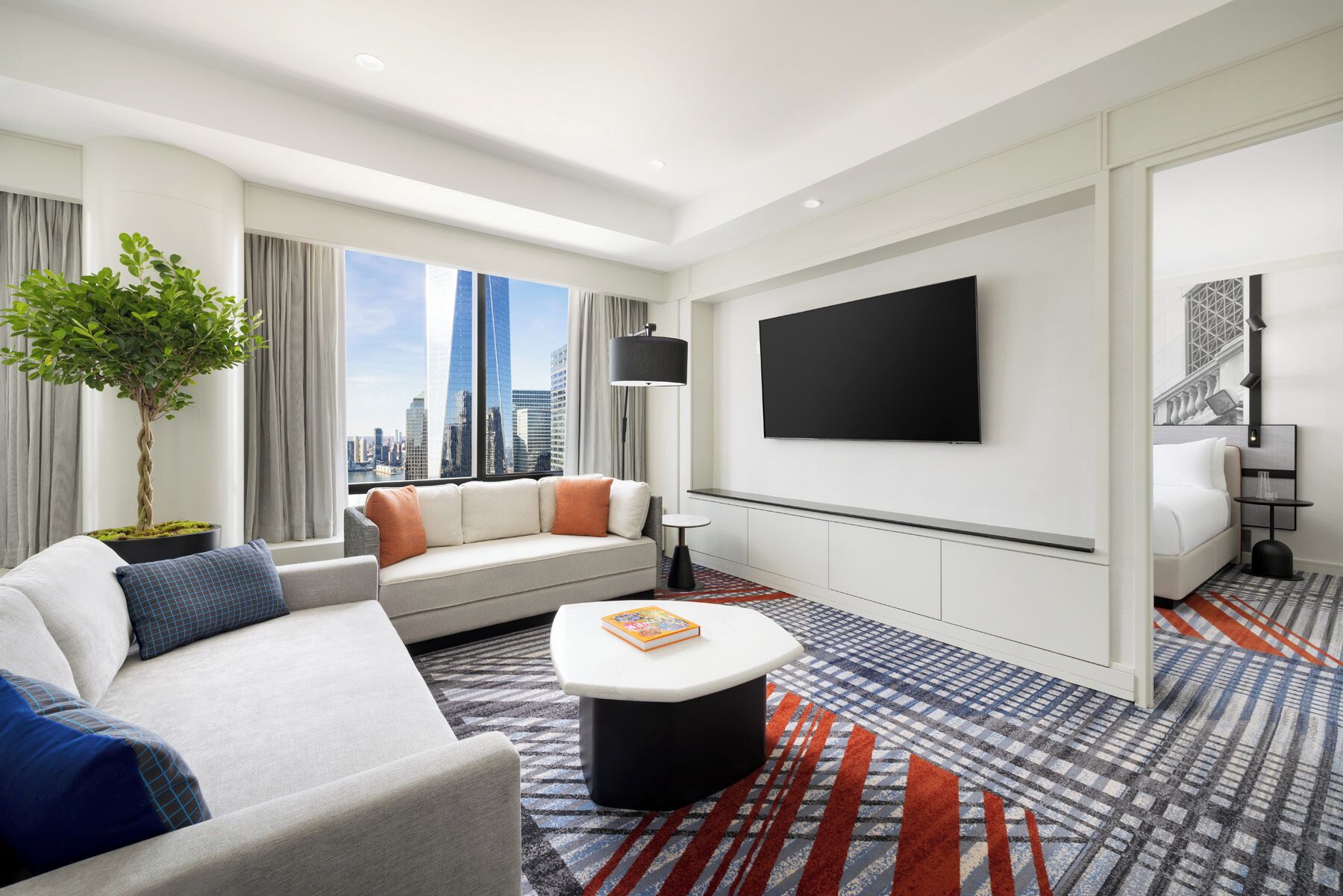 Quarto de hotel moderno com sofá, mesa de centro, TV de tela plana, planta em vaso e vista para a cidade.