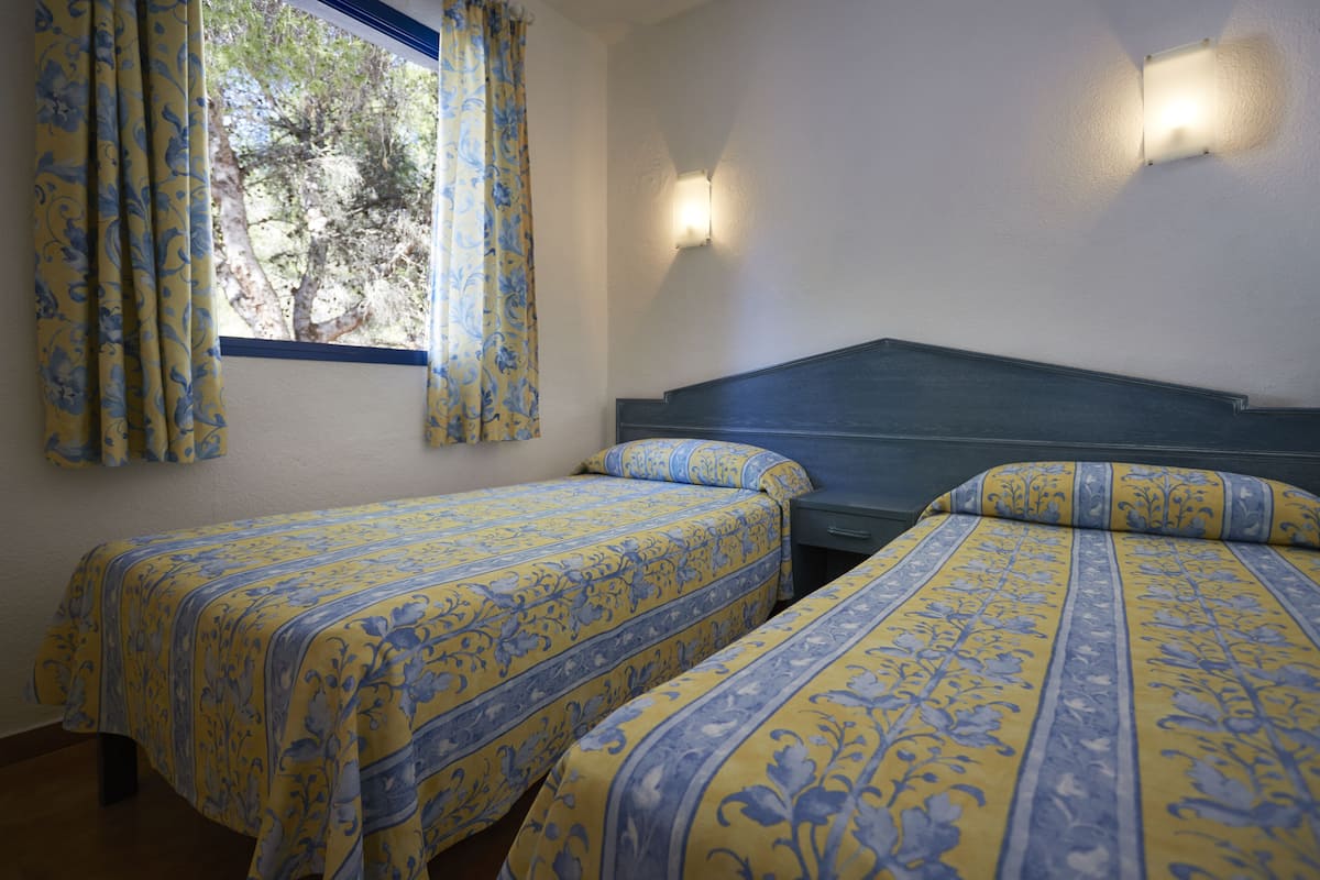 Quarto com duas camas, cada uma com colcha estampada, uma janela com cortinas e luminárias na parede.