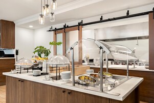 Buffet montado com cubas de vidro transparente, travessas de aço inoxidável e área de cozinha moderna ao fundo.