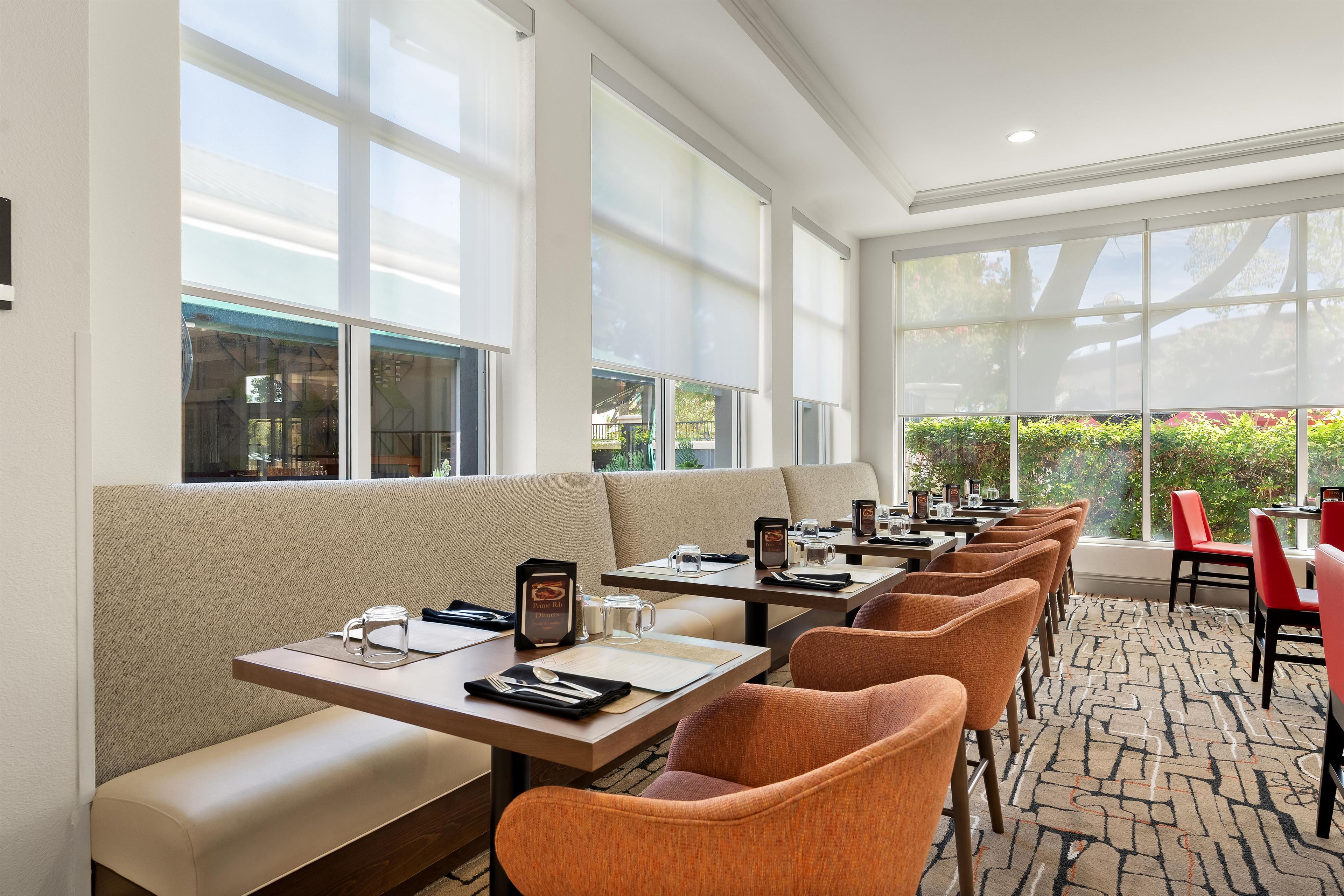 Restaurante com tetos altos, grandes janelas e área de estar com bancos estofados e cadeiras laranja.