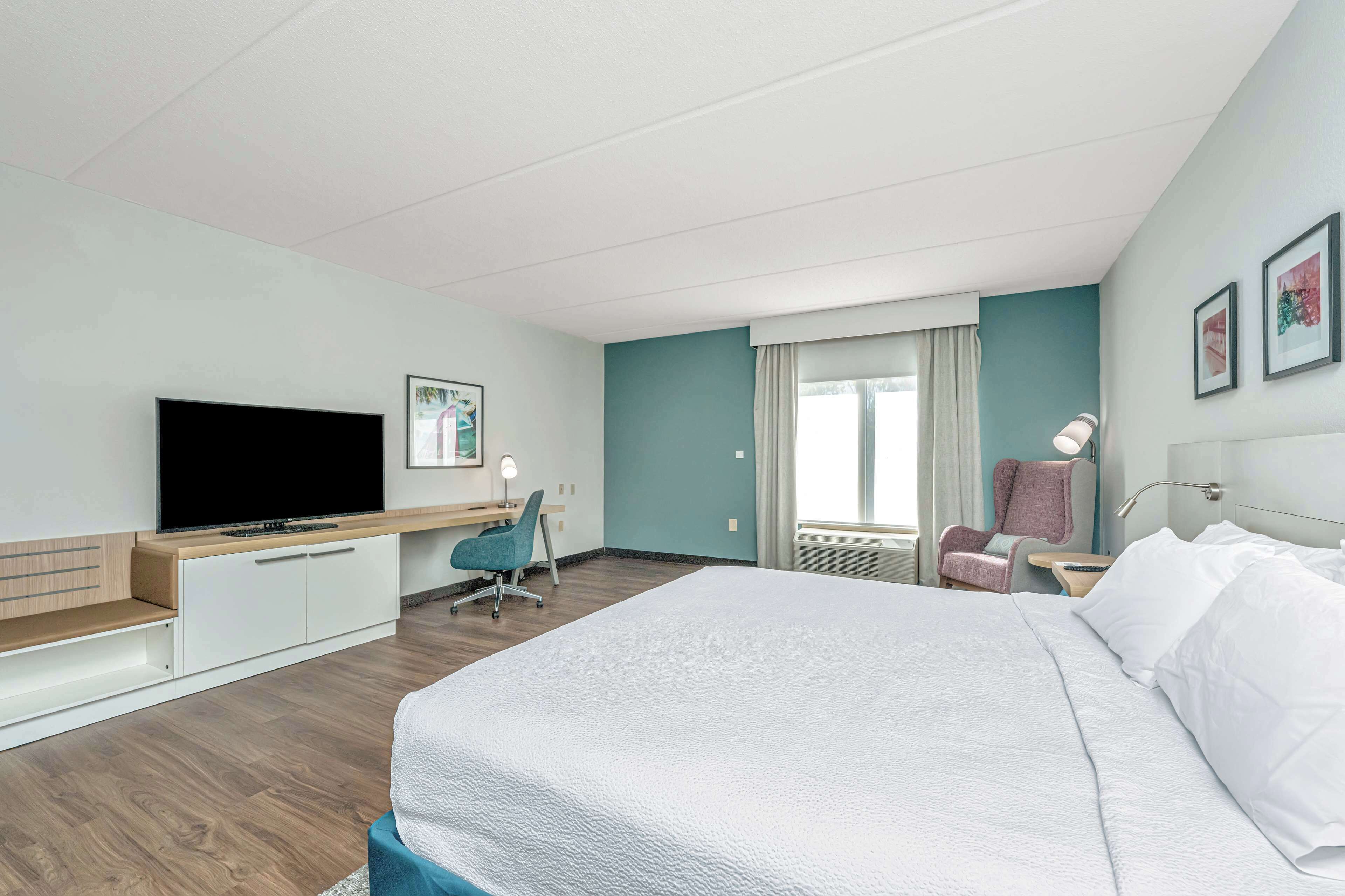 Quarto de hotel com uma cama grande, uma escrivaninha com computador, televisão e janela com cortinas.