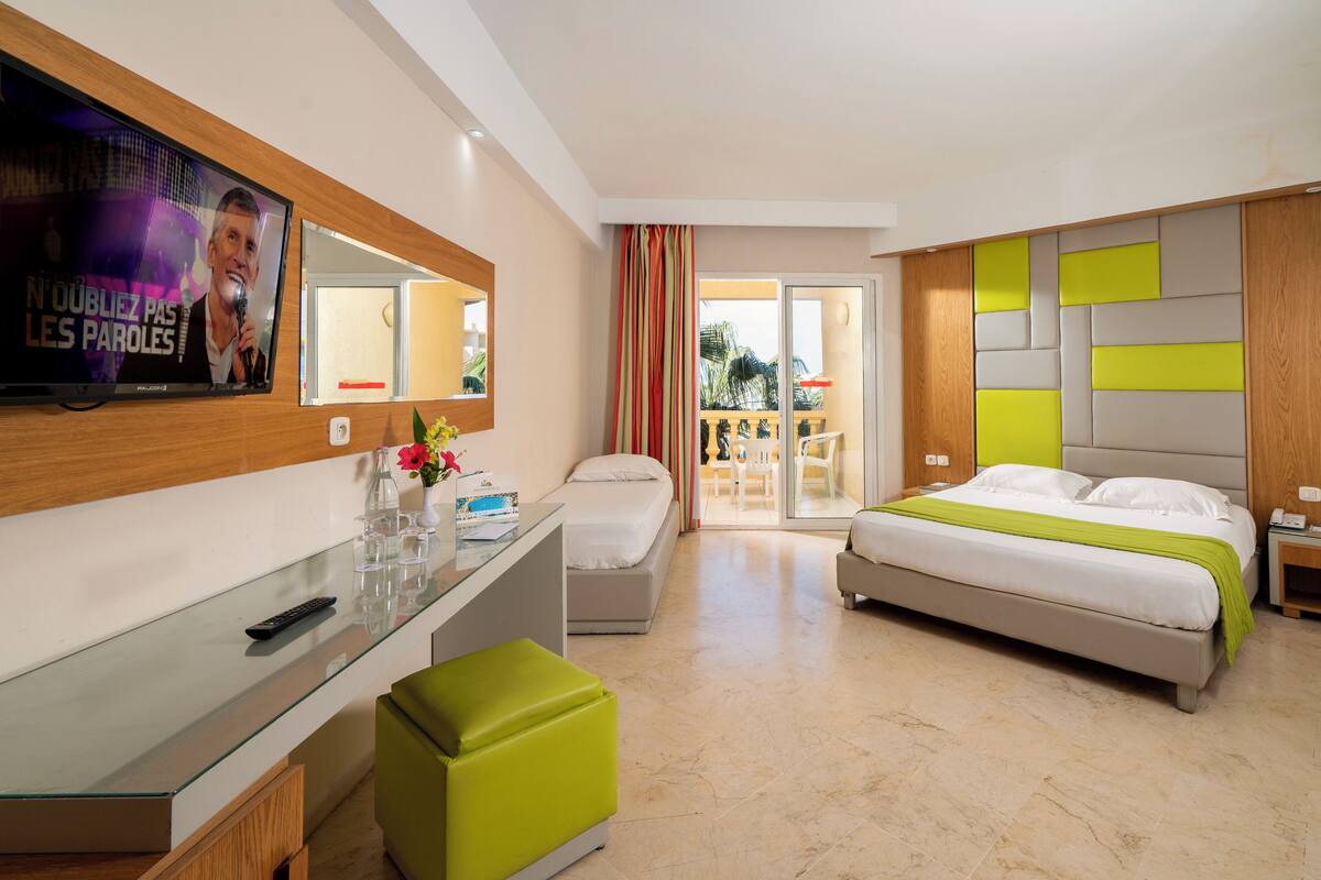 Quarto de hotel com cama, escrivaninha com espelho, televisão, varanda com vista e janela com cortinas.