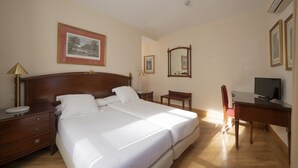 Quarto de hotel bem organizado, com uma cama grande, mesas de cabeceira, escrivaninha com cadeira, televisão e quadros nas paredes.