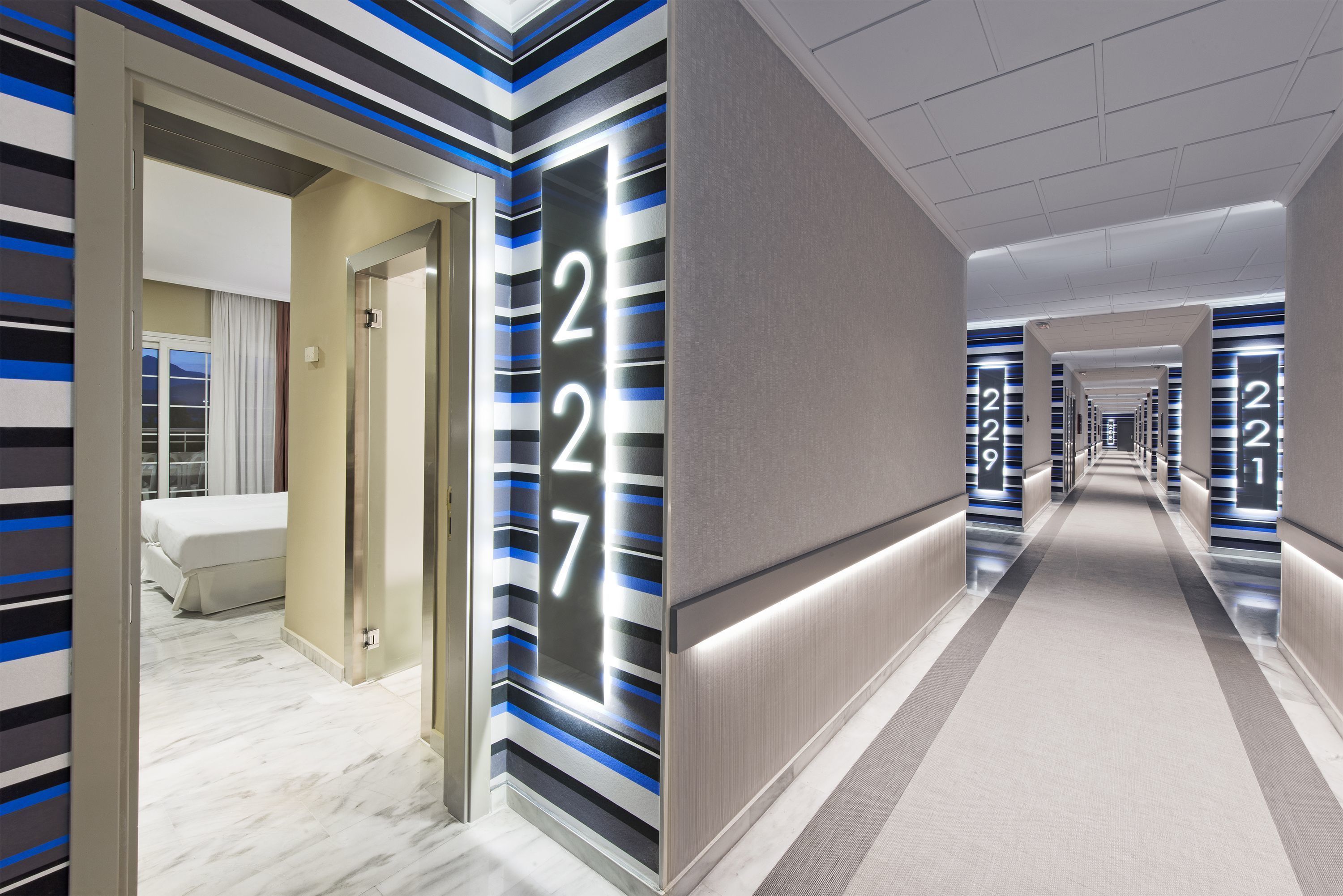 Corredor de hotel com design moderno, exibindo um número na parede e piso coberto de carpete.