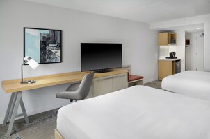 Quarto de hotel com uma cama, uma escrivaninha com um computador, uma televisão, uma cadeira e uma cozinha compacta.
