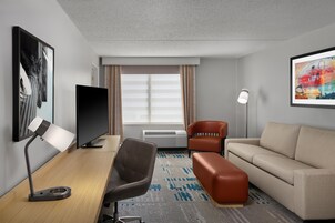 Quarto de hotel com escrivaninha, cadeira, sofá, televisão, aparelho de ar condicionado e obras de arte nas paredes.