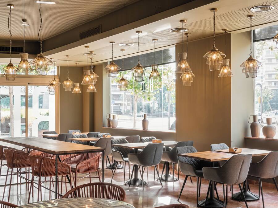 Um café amplo com várias mesas e cadeiras, luminárias pendentes e grandes janelas que oferecem vista para a rua.