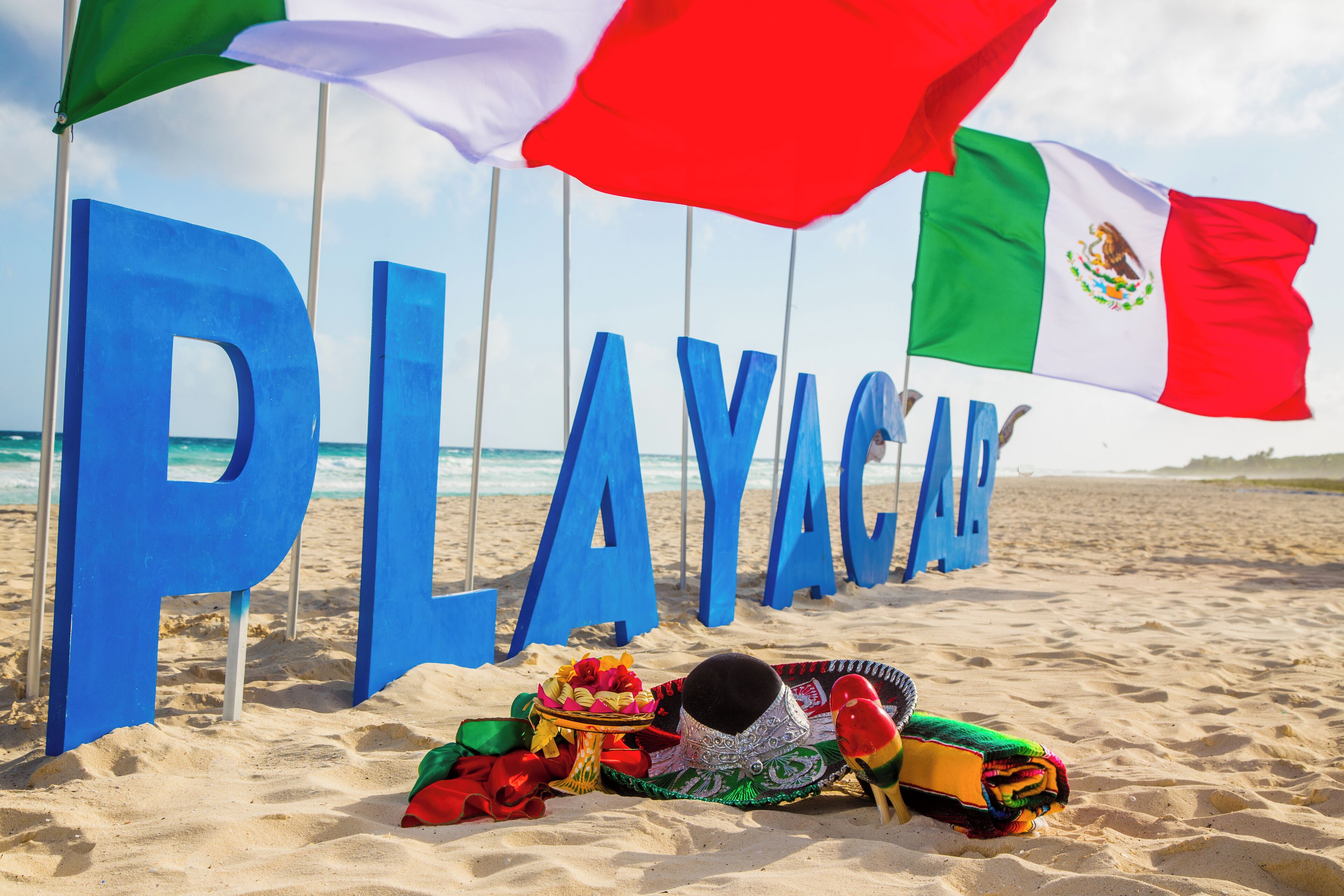 Uma praia com letras azuis grandes formando a palavra 'PLAYACU' e uma bandeira mexicana.