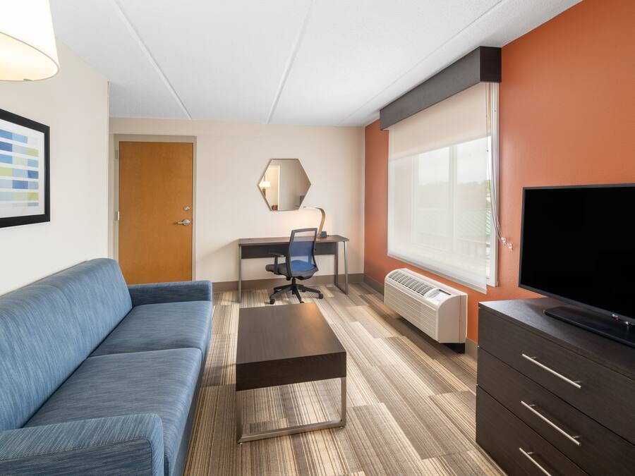 Quarto de hotel moderno com um sofá azul, uma escrivaninha com cadeira, televisão e uma janela grande.