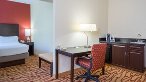 Quarto de hotel com cama, escrivaninha, cadeira, luminária e cozinha compacta.