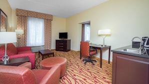 Quarto de hotel com sofá vermelho, uma mesa, uma cadeira, televisão, luminária e pia.