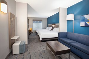 Quarto de hotel com cama, sofá, escrivaninha e cadeira. Há uma janela com persianas e um abajur na parede.