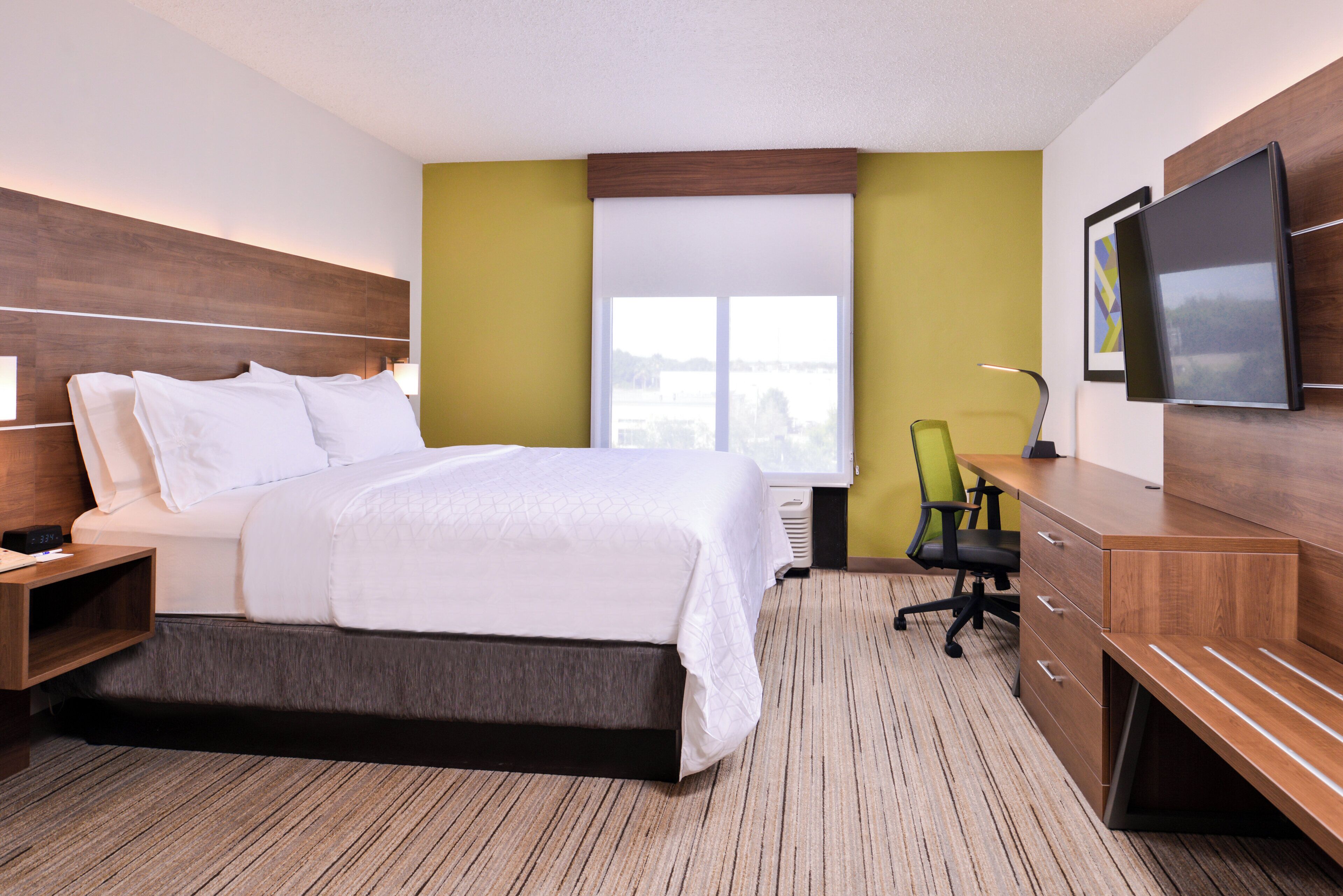 Quarto de hotel com cama, escrivaninha, cadeira, televisão e janela.