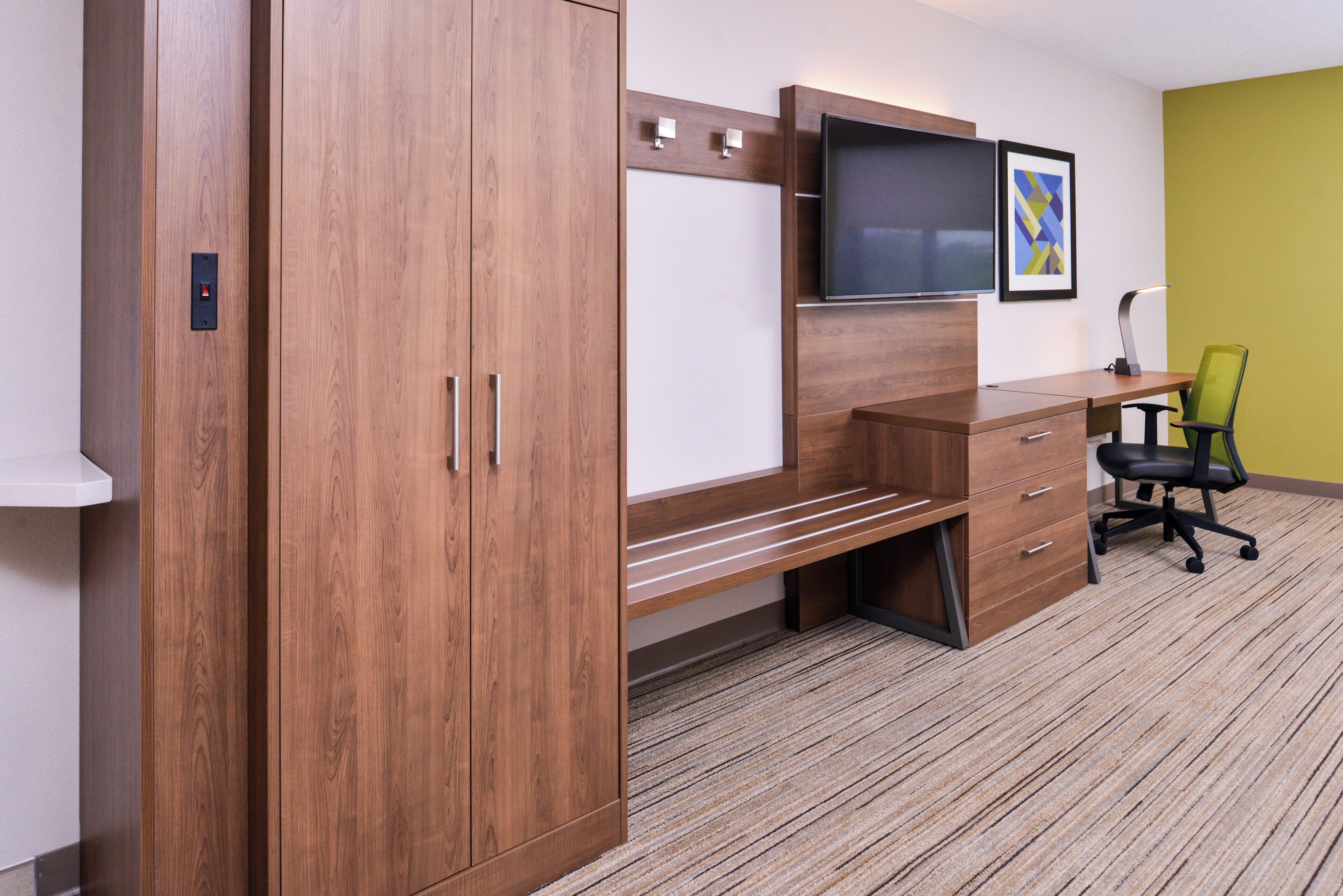 Quarto de hotel com escrivaninha de madeira, TV de tela plana, cadeira verde e guarda-roupa espaçoso.