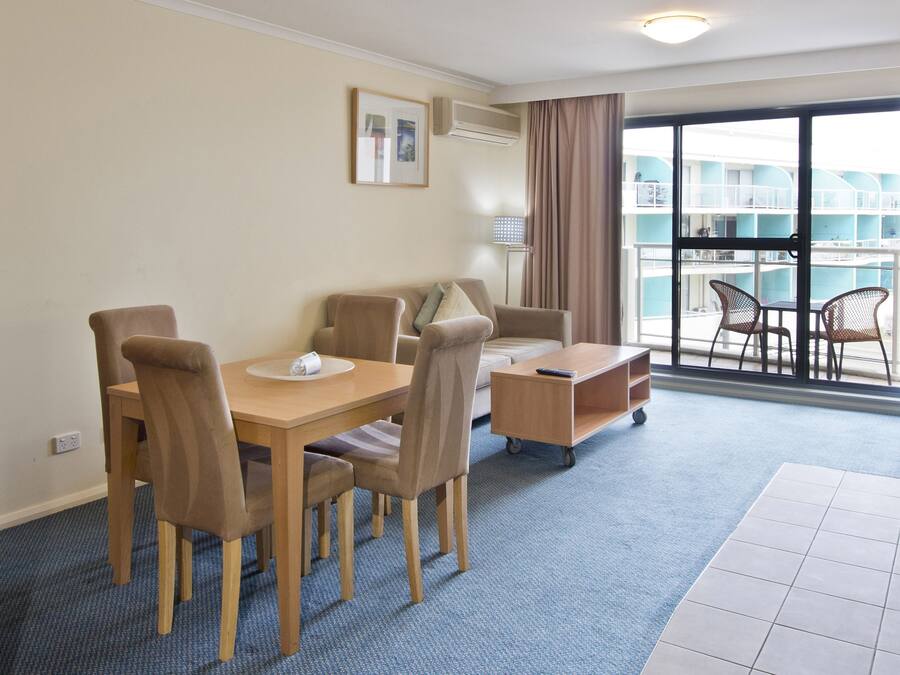 Quarto de hotel com mesa de jantar, cadeiras, um pequeno sofá, mesa lateral e vista para uma piscina e um edifício através de grandes janelas.