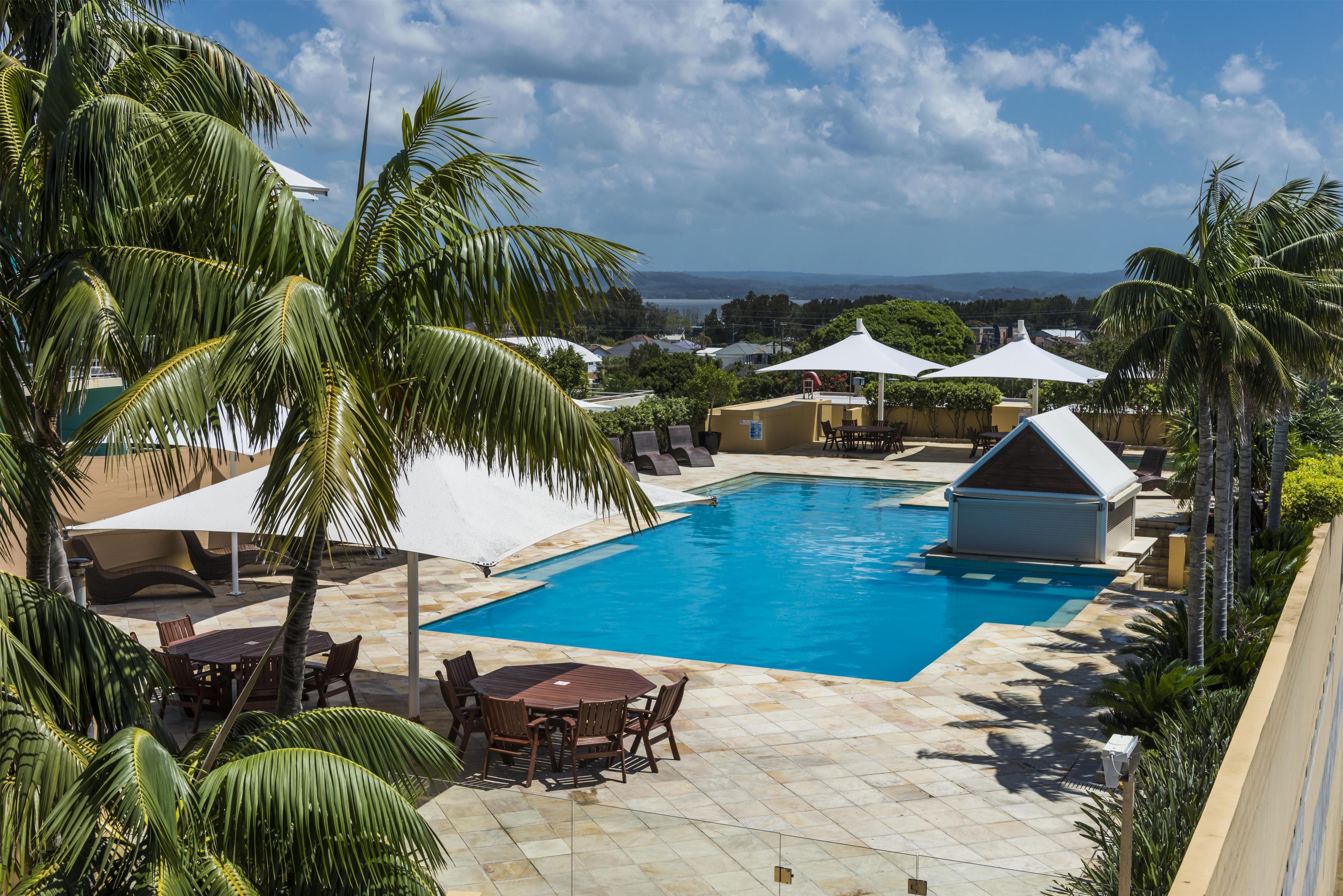 Área de piscina com vista para o oceano, cercada por palmeiras e assentos cobertos.
