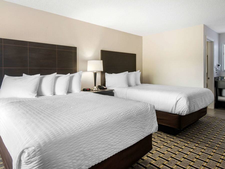 Quarto de hotel com duas camas, uma mesa de cabeceira com abajur, banheiro com janela e piso estampado.