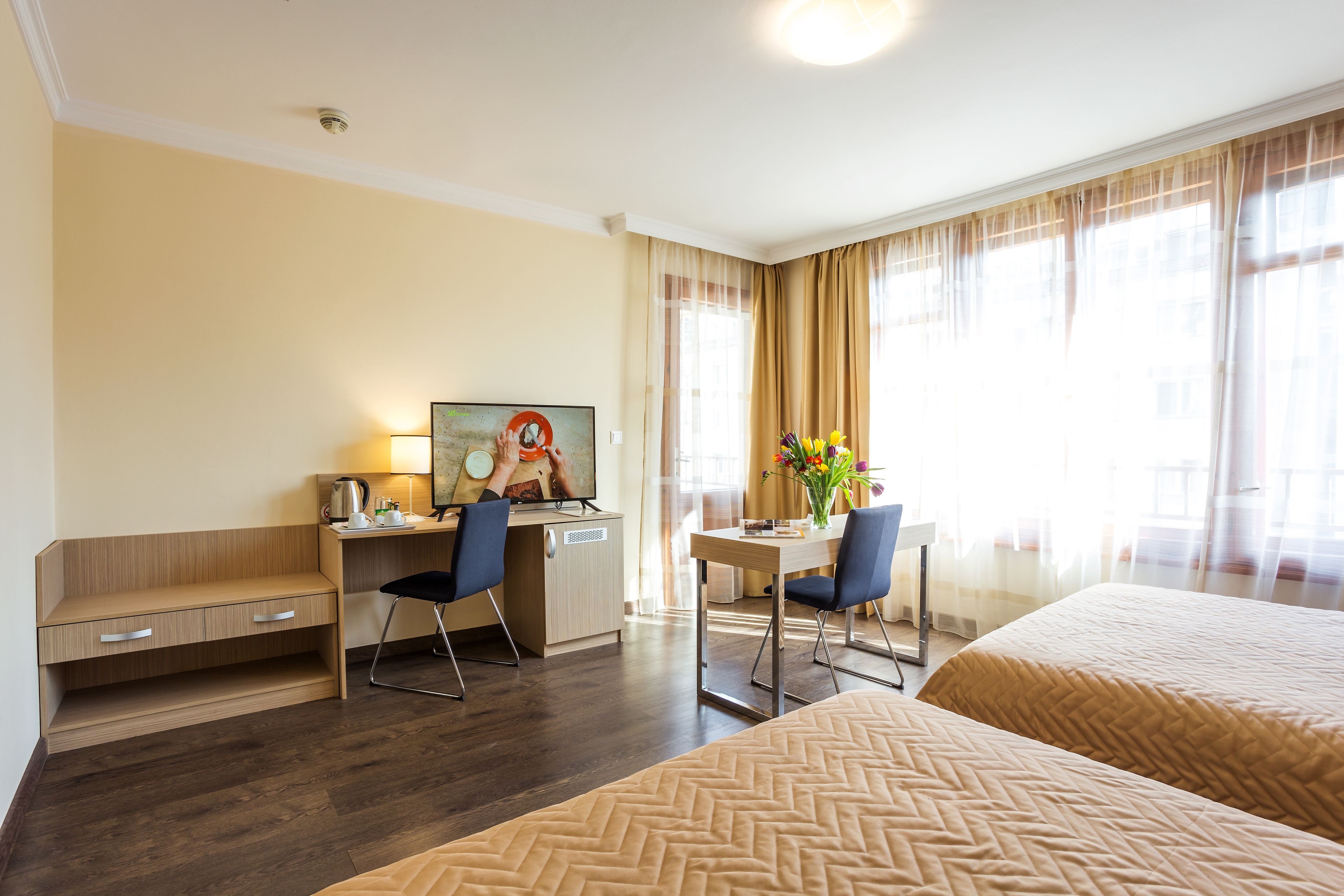 Quarto de hotel com cama, escrivaninha com computador, cadeira, mesa pequena com um vaso de flores e janela com cortinas.