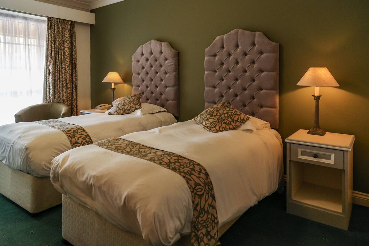 Quarto de hotel com duas camas, cada uma com cabeceira estofada, mesa de cabeceira com abajur e uma cadeira.
